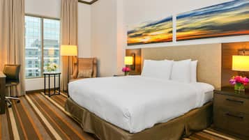 Quarto de hotel com uma cama grande, uma escrivaninha, uma cadeira, uma mesinha e uma janela com cortinas.