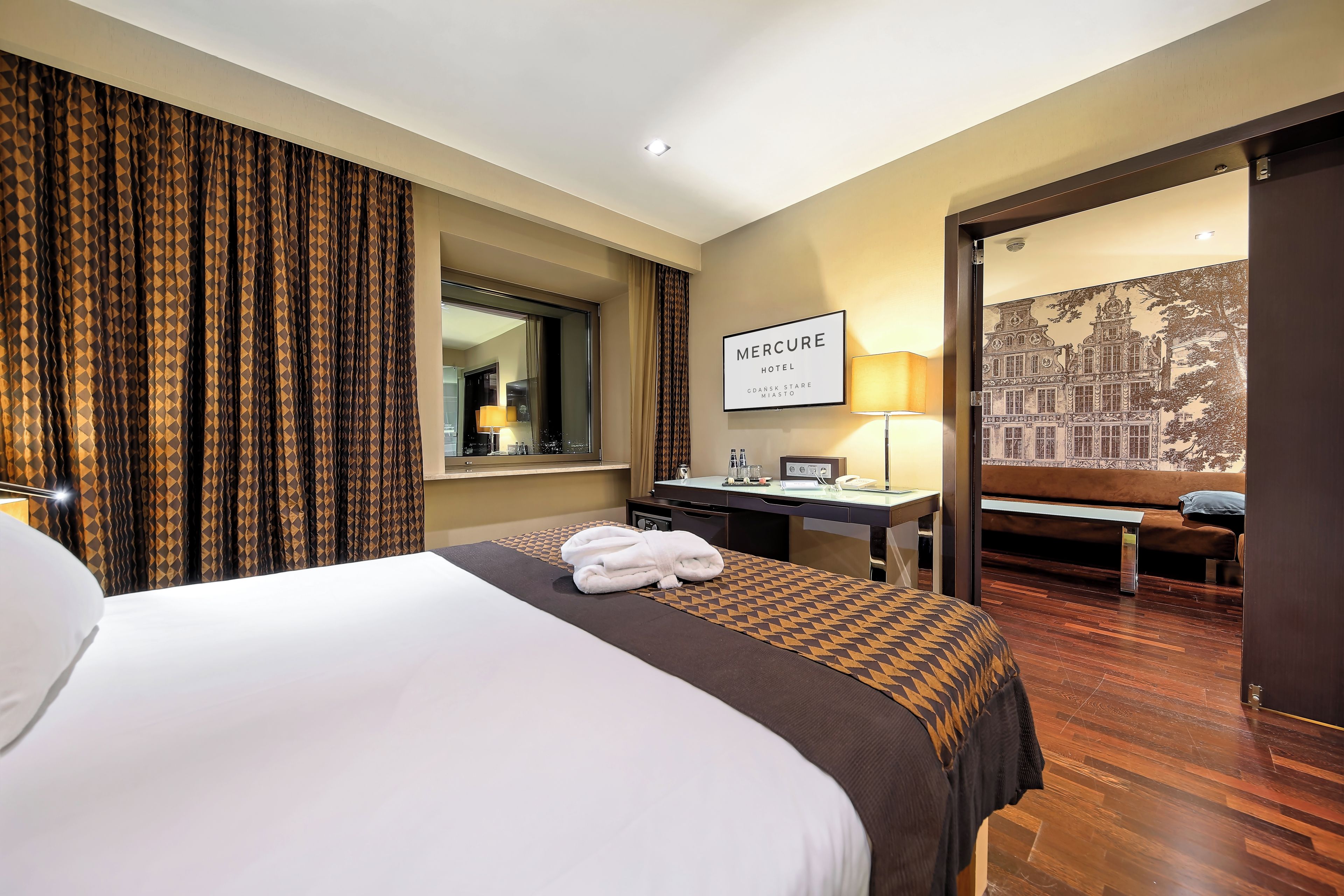Quarto de hotel com cama, escrivaninha, espelho, placa na parede e vista de um edifício através de uma porta aberta.