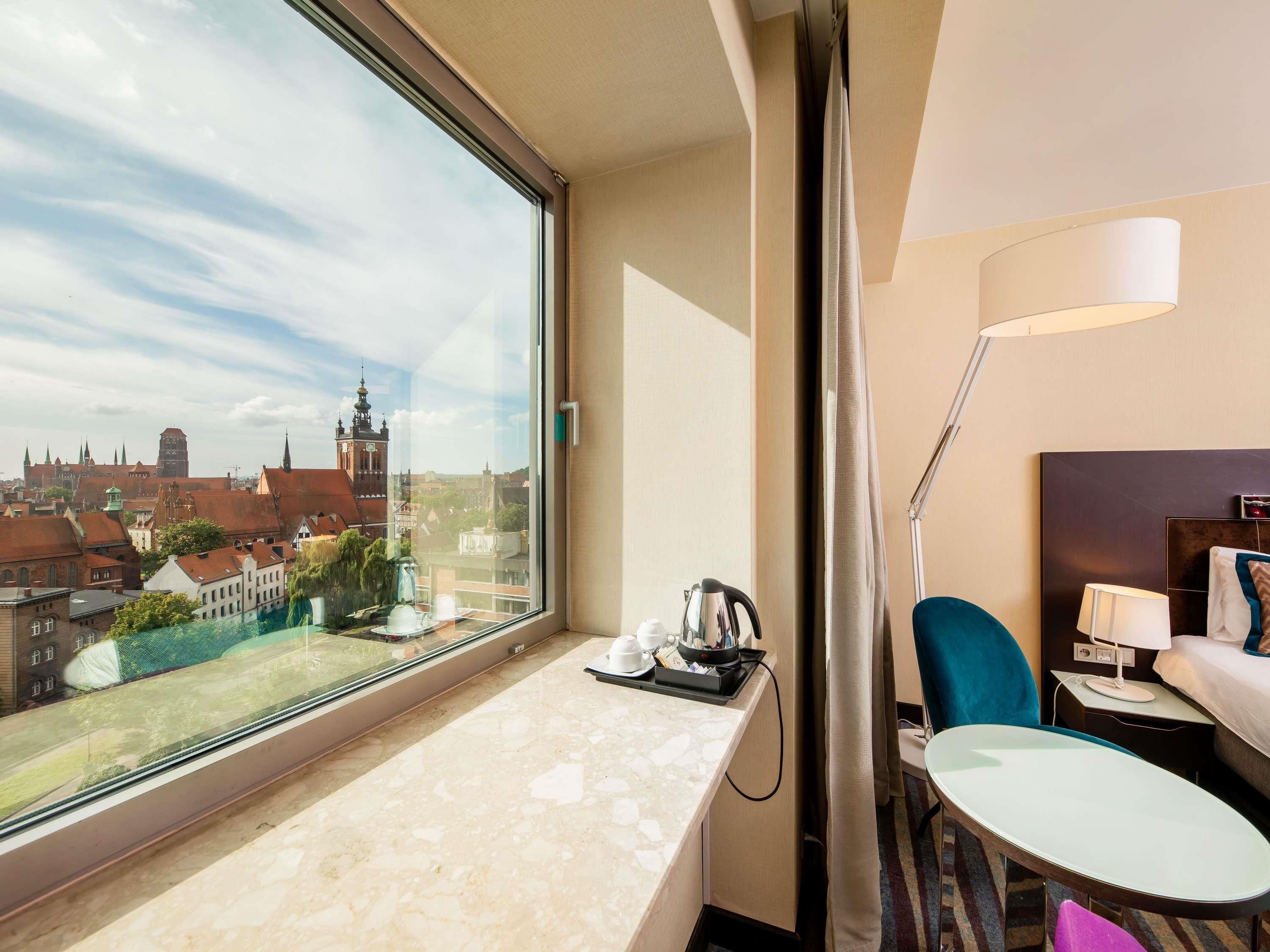 Quarto de hotel com uma janela grande, uma mesa, uma cadeira, um abajur e uma cama.