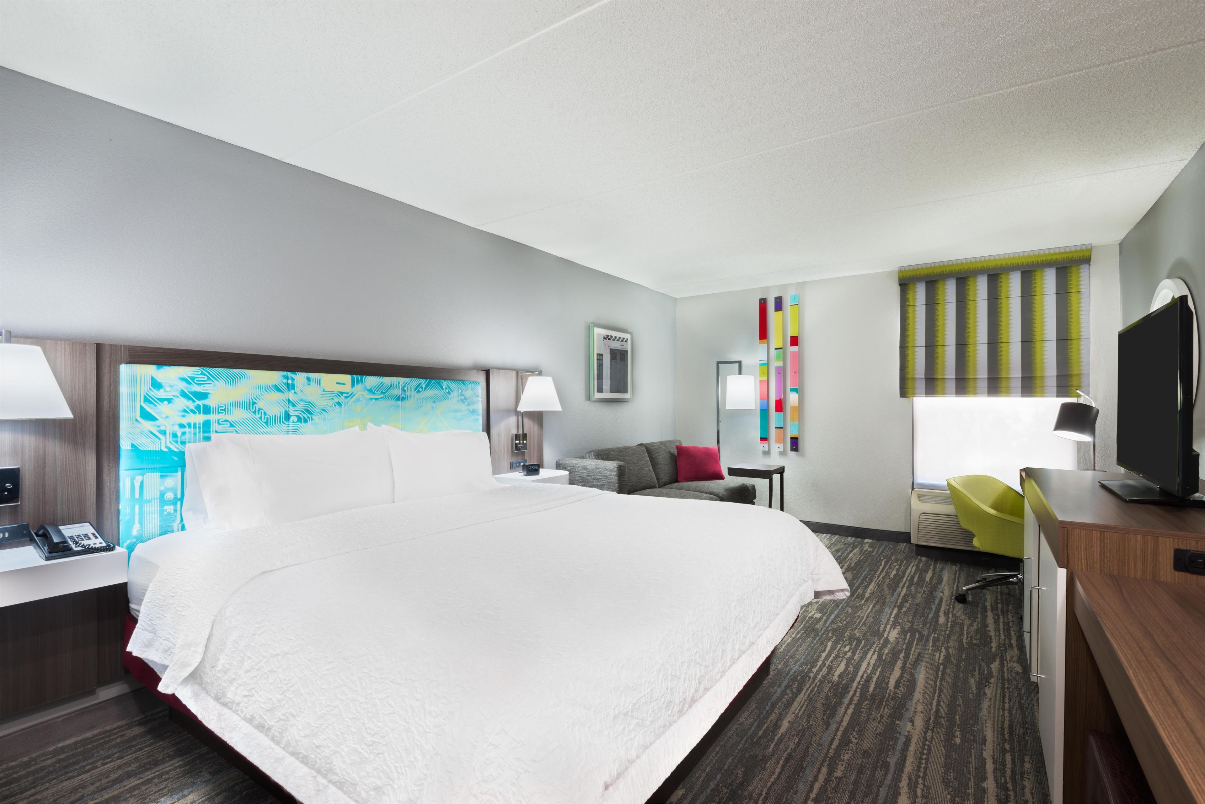 Quarto de hotel com uma cama grande, uma escrivaninha com computador, uma cadeira, televisão e janela com persianas.