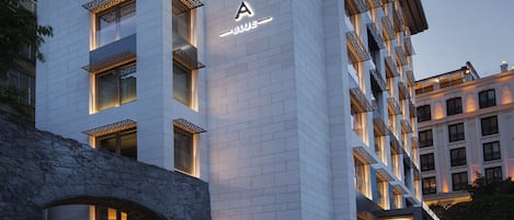 Um hotel moderno com uma placa de destaque que diz "Hotel Arcadia Blue". Apresenta várias varandas com iluminação, uma entrada em arco de pedra e uma fachada bem iluminada.