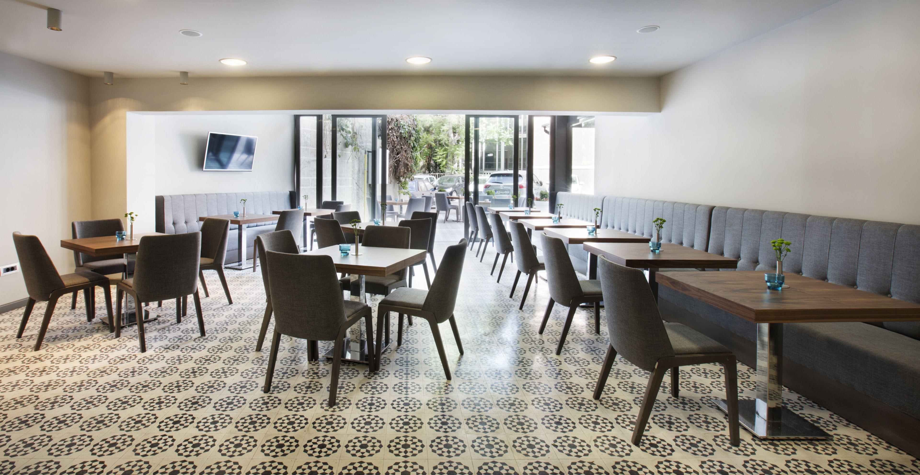 Um restaurante com chão estampado, várias mesas e cadeiras, e uma grande janela com vista para a rua.