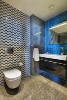 Uma casa de banho moderna com parede revestida de azulejos, sanita, lavatório e espelho.