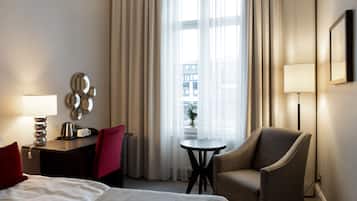 Quarto de hotel com cama, escrivaninha, cadeira, mesa e janela com cortinas.