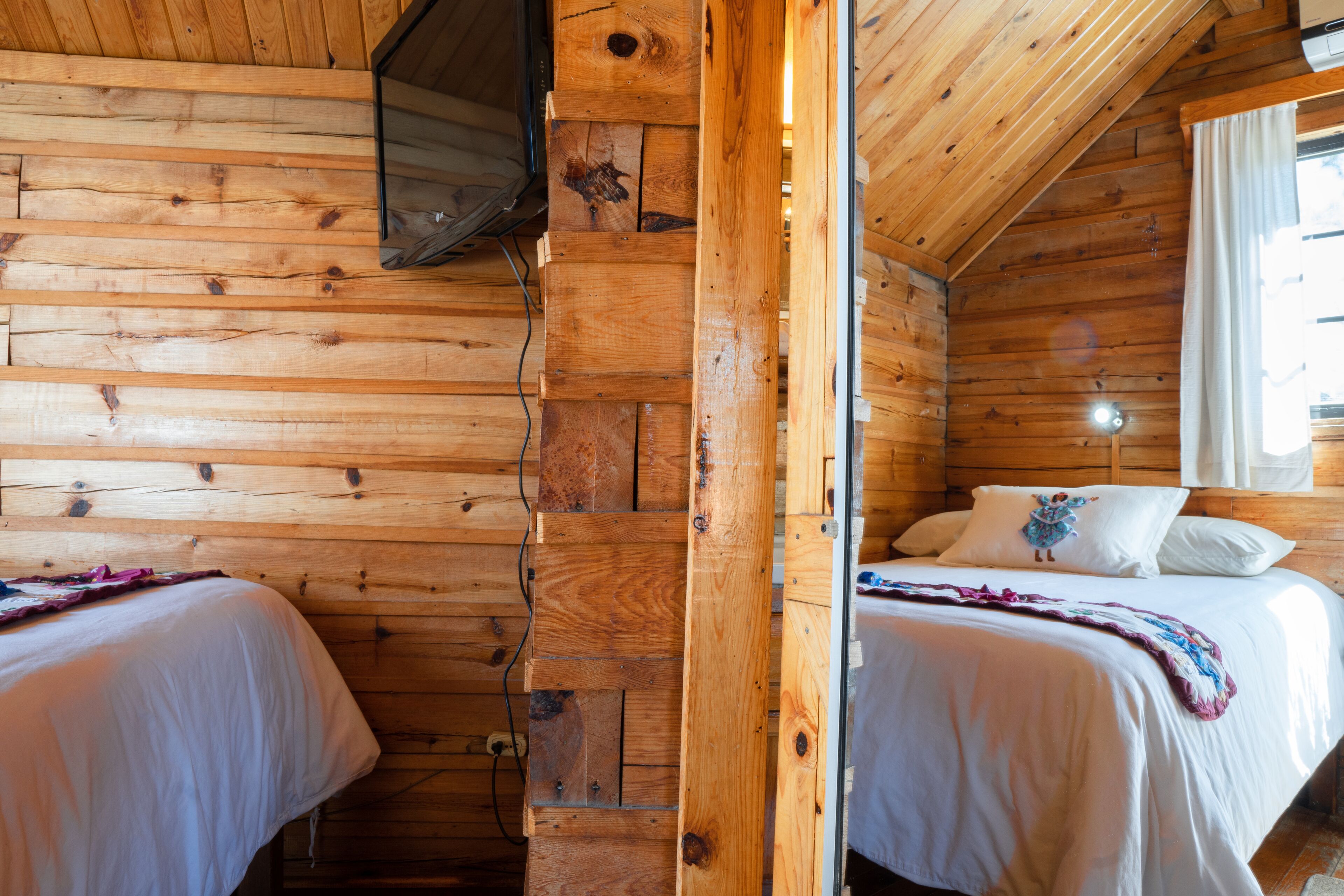 Quarto de cabana de madeira com duas camas, teto de madeira e janela com cortinas.