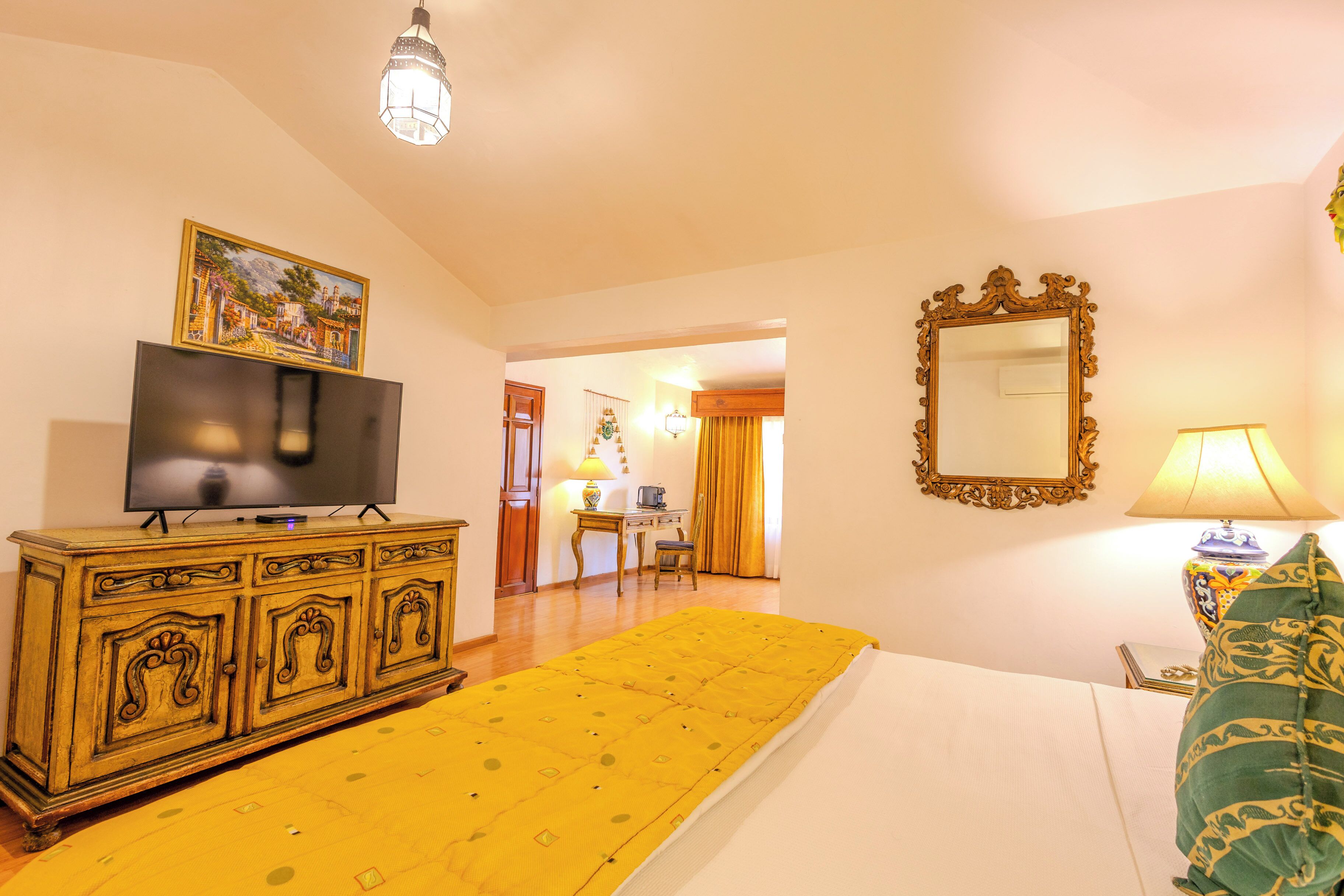 Quarto de hotel com cama, televisão, penteadeira, espelho e um quadro na parede.
