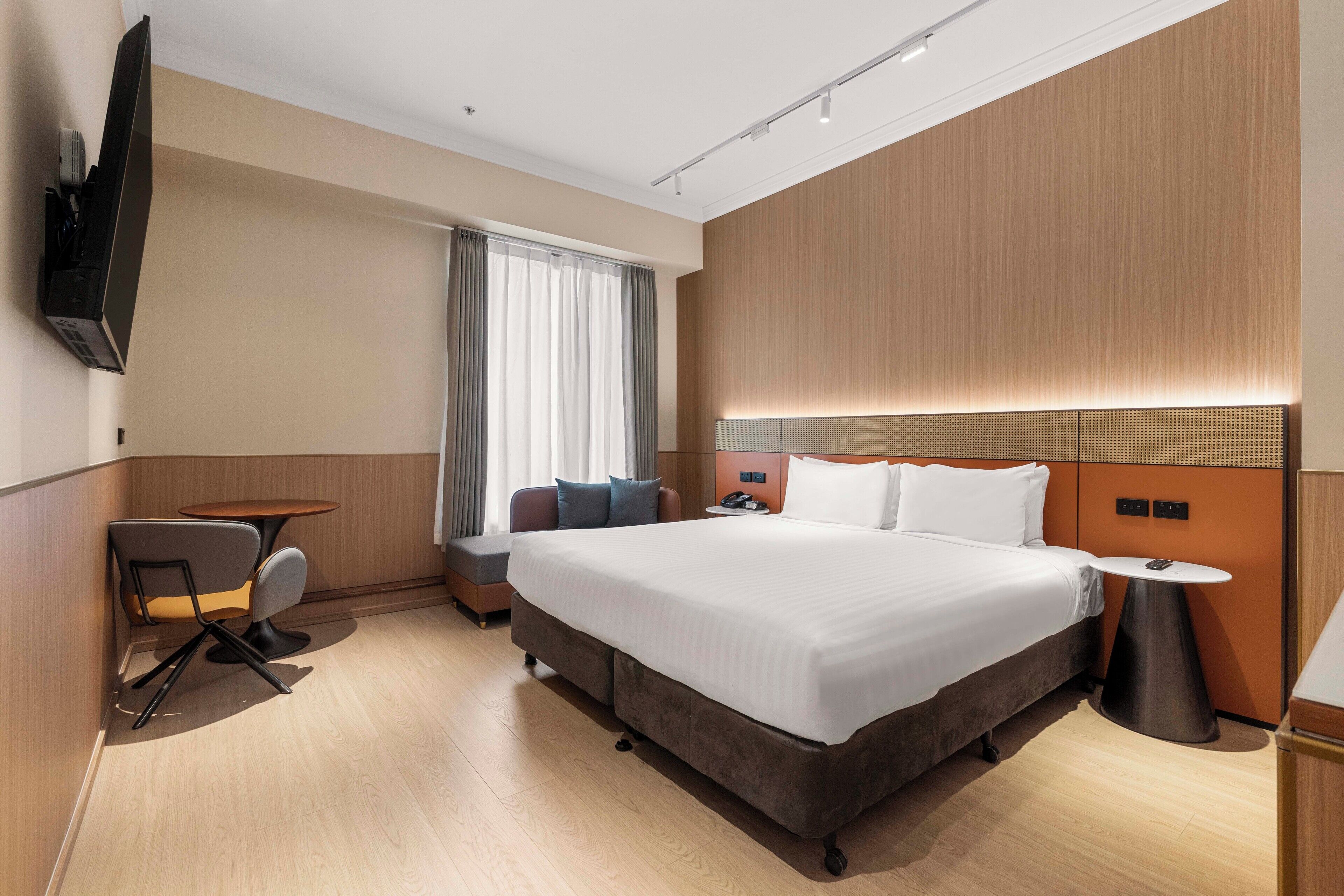Quarto de hotel moderno com uma cama grande, uma escrivaninha, uma cadeira, uma TV fixada na parede e uma janela com cortinas.