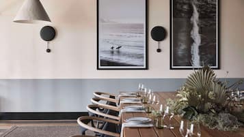Uma sala de jantar com uma longa mesa de madeira posta para seis pessoas, com dois alto-falantes pretos fixados na parede e obras de arte oceânicas emolduradas.