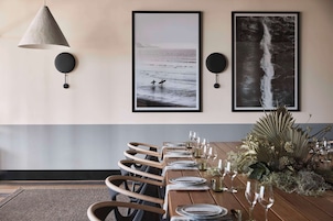 Uma sala de jantar com uma longa mesa de madeira posta para seis pessoas, com dois alto-falantes pretos fixados na parede e obras de arte oceânicas emolduradas.