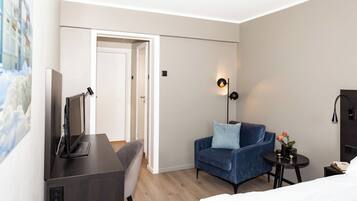 Quarto de hotel moderno com cama, escrivaninha com computador, poltrona azul, pequena mesa com velas e luminária na parede.