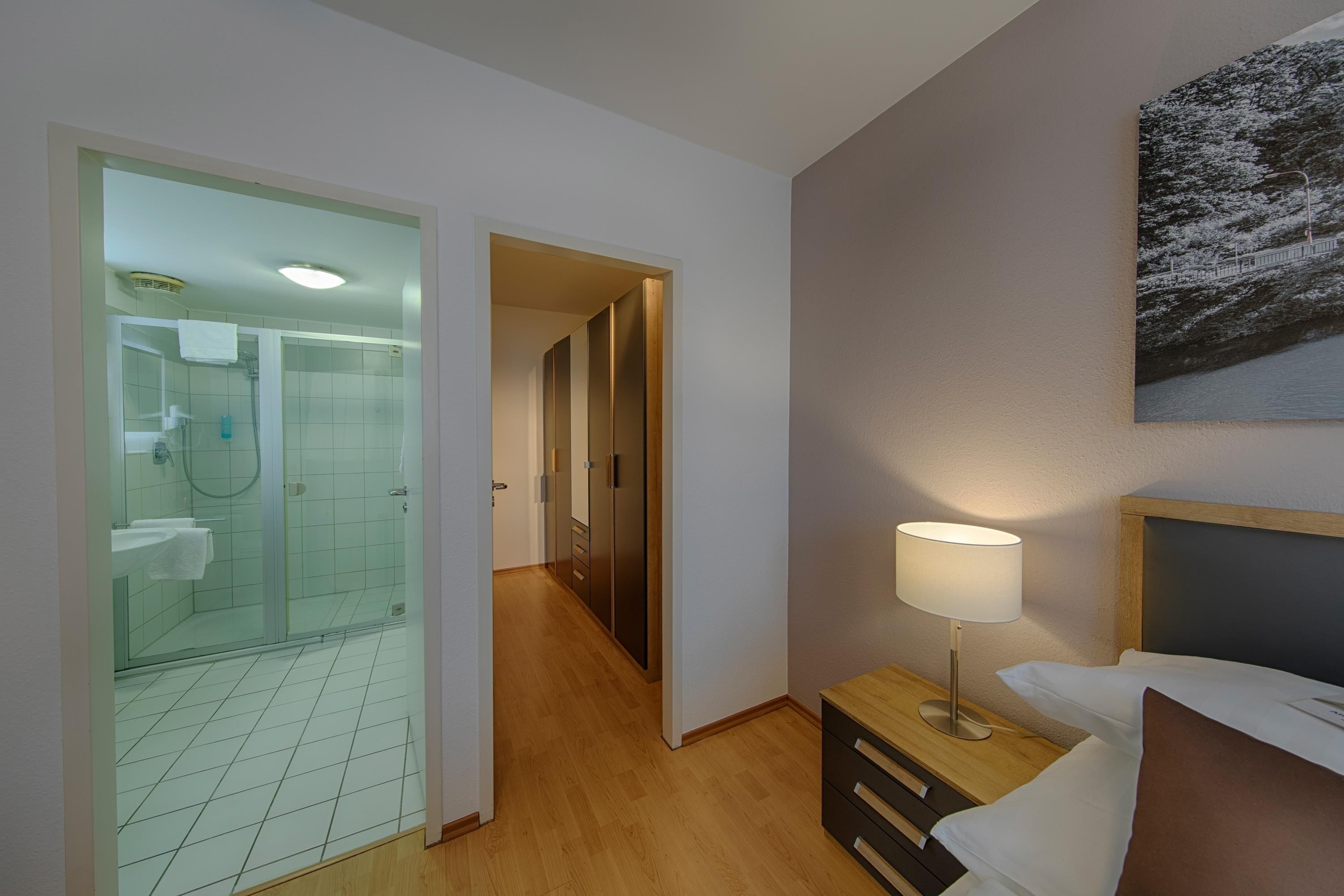 Quarto de hotel com chuveiro em box de vidro, mesa de cabeceira com abajur e um quadro na parede.