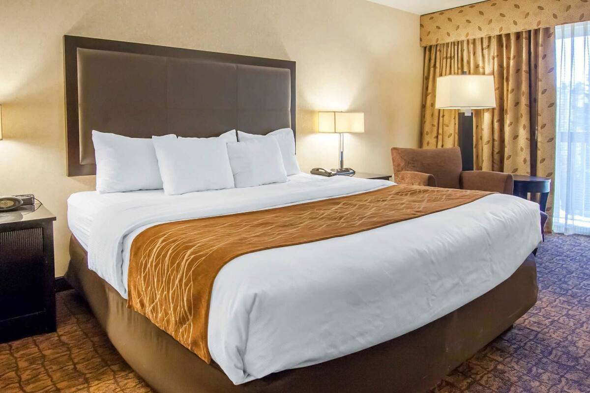 Quarto de hotel com uma cama grande, uma cadeira, uma escrivaninha com abajur e uma janela com cortinas.