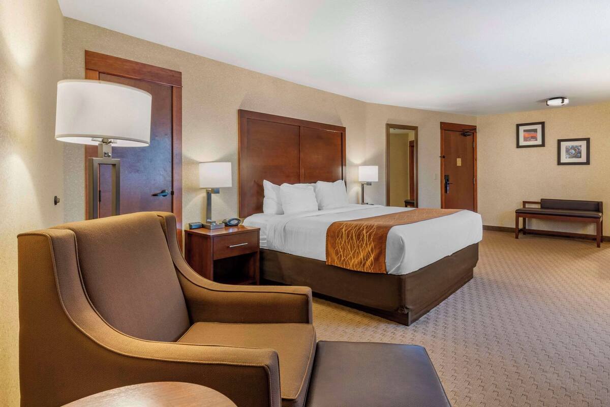Quarto de hotel com uma cama grande, duas mesas de cabeceira com luminárias, uma poltrona marrom e uma mesa pequena.