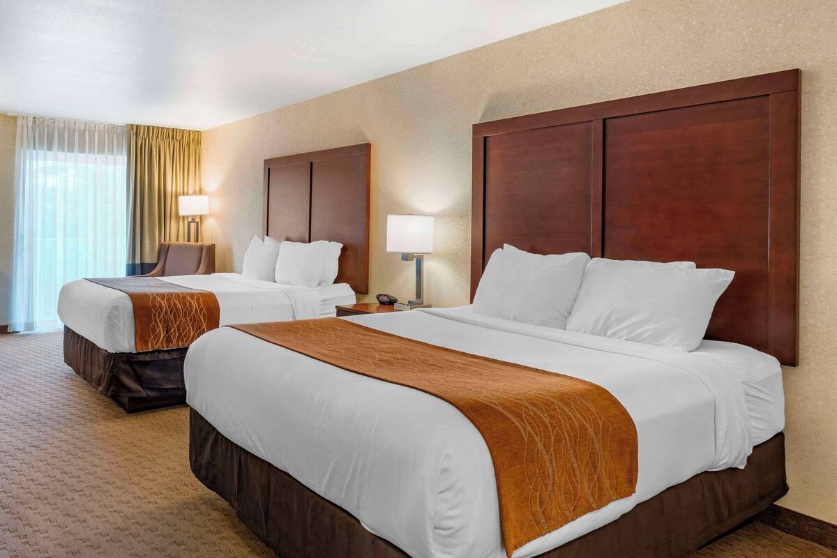 Quarto de hotel com duas camas, uma cabeceira de madeira grande, mesa de cabeceira com abajur e vista para o exterior através de uma janela com cortinas.