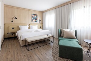 Quarto de hotel com cama, poltrona verde, mesa pequena e um quadro na parede.