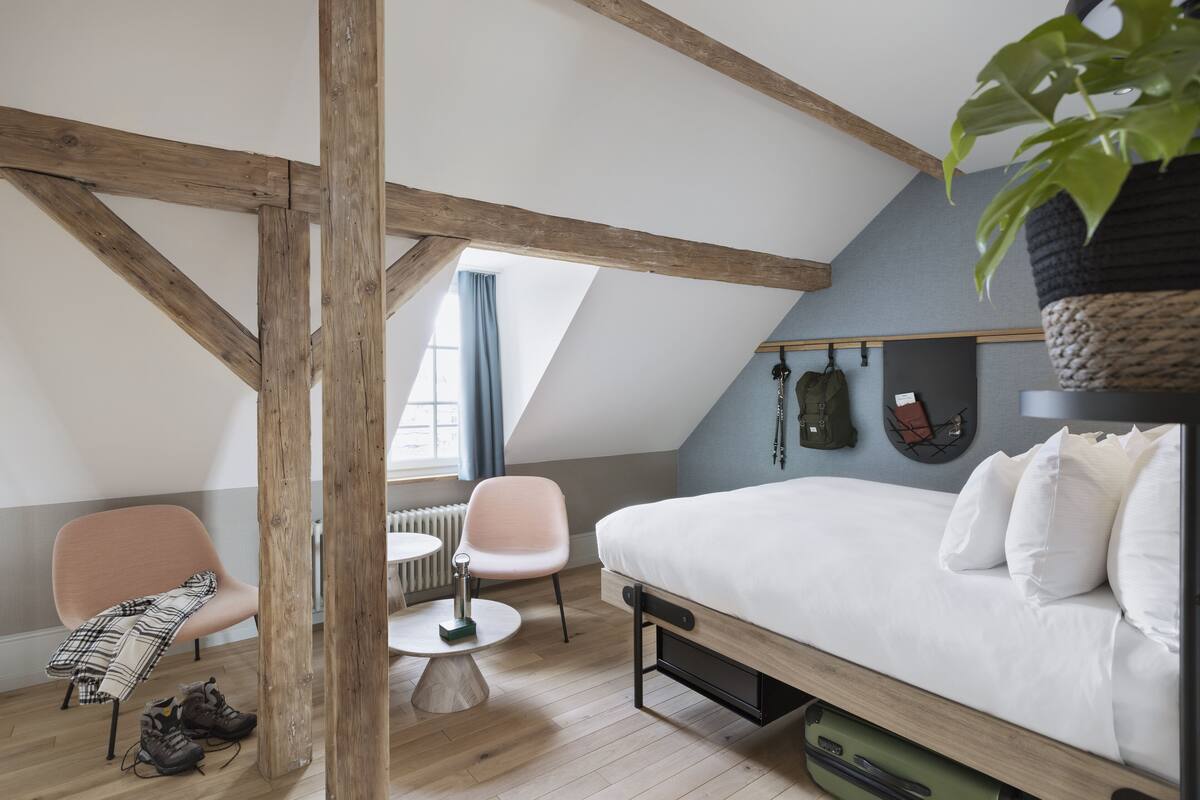 Quarto com teto inclinado, vigas de madeira, cama, cadeira, mesa pequena e uma planta.