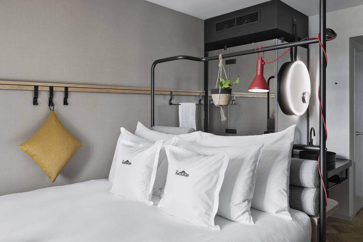 Cama bem arrumada com roupa de cama e almofadas brancas, uma planta pendente, um abajur vermelho e uma coluna de som preta sobre um suporte de metal.