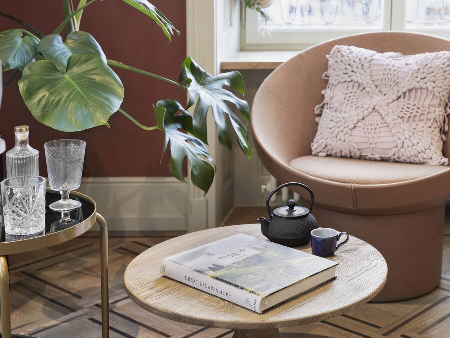 Um recanto aconchegante para leitura, com uma mesa redonda de madeira, uma poltrona confortável com almofada tricotada, um livro intitulado 'Great Escapes Alps', um bule e taças decorativas.