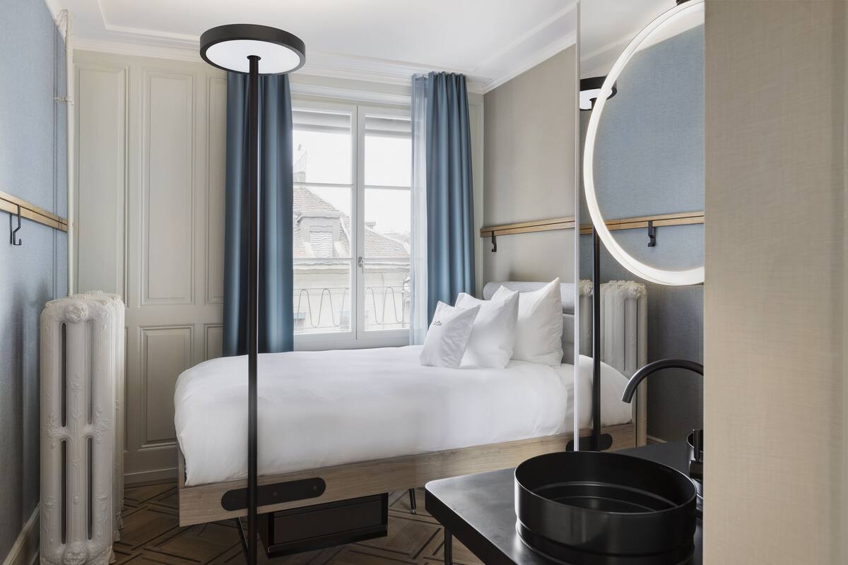 Quarto de hotel com cama, espelho redondo, mesa de cabeceira e janela com cortinas.