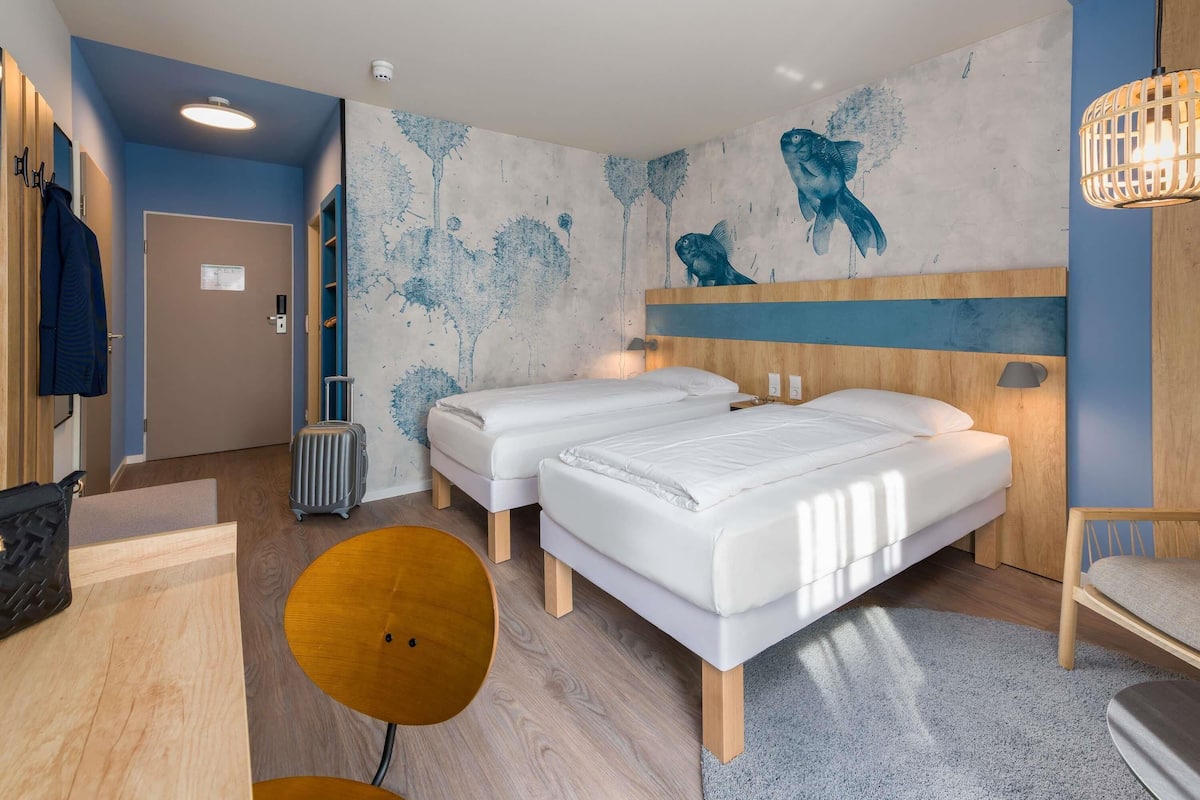 Quarto de hotel moderno com duas camas, cabeceira de madeira, painel na parede, mesa de madeira redonda e radiador.