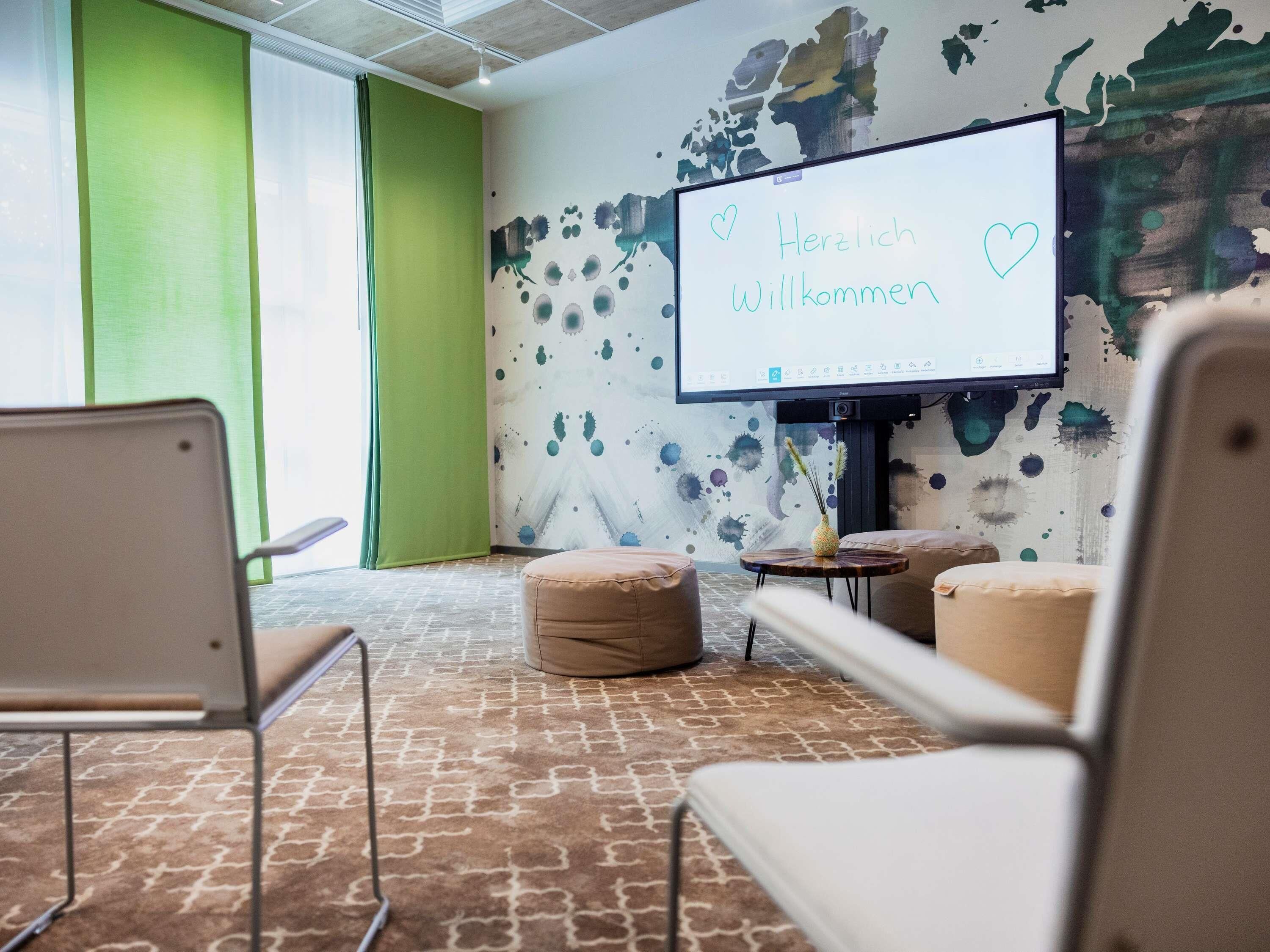 Uma sala de conferências com uma tela de projetor exibindo 'Herzlich Willkommen' e um símbolo de coração, uma parede verde, um carpete com padrão de tijolos e duas poltronas estofadas.