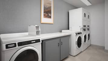 Uma lavanderia com máquina de lavar comercial Maytag, armários cinzas e um quadro com paisagem na parede.