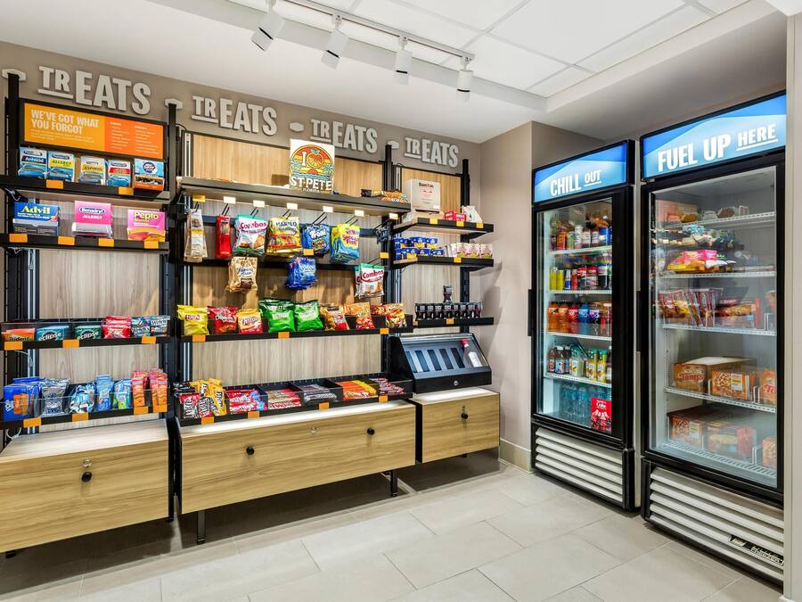 Espaço para lanches e bebidas com autosserviço, com diversas opções de snacks embalados, um caixa registradora e dispensadores refrigerados de bebidas.