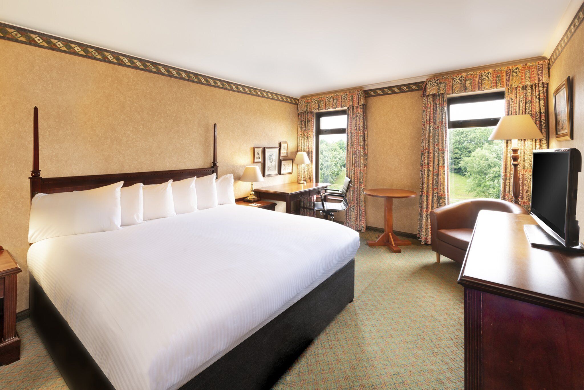 Quarto de hotel com uma cama grande, uma mesa com cadeira, uma televisão e uma janela com cortinas.
