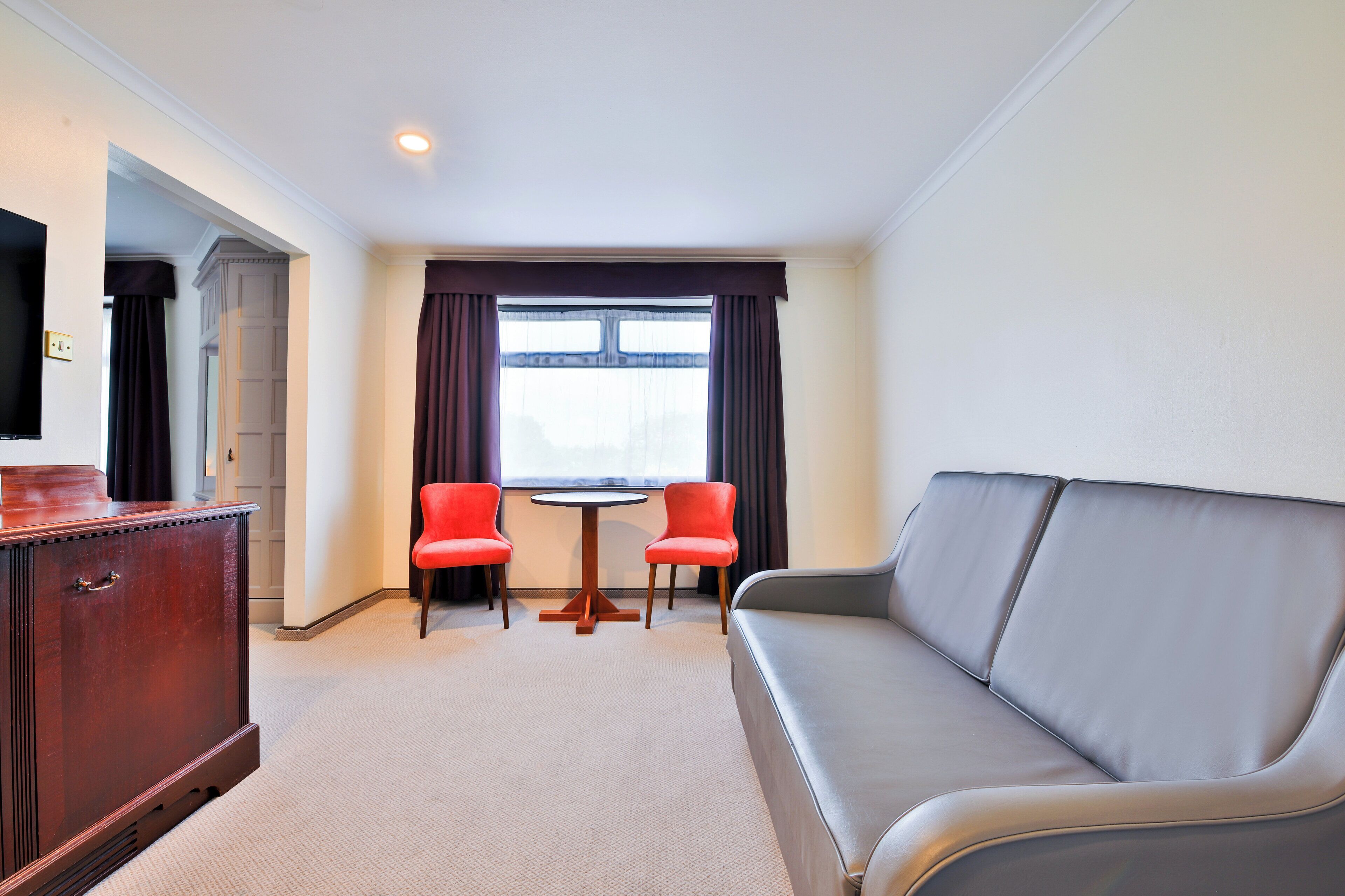 Quarto de hotel com um sofá cinzento, duas cadeiras vermelhas, um armário de madeira e uma pequena mesa redonda.