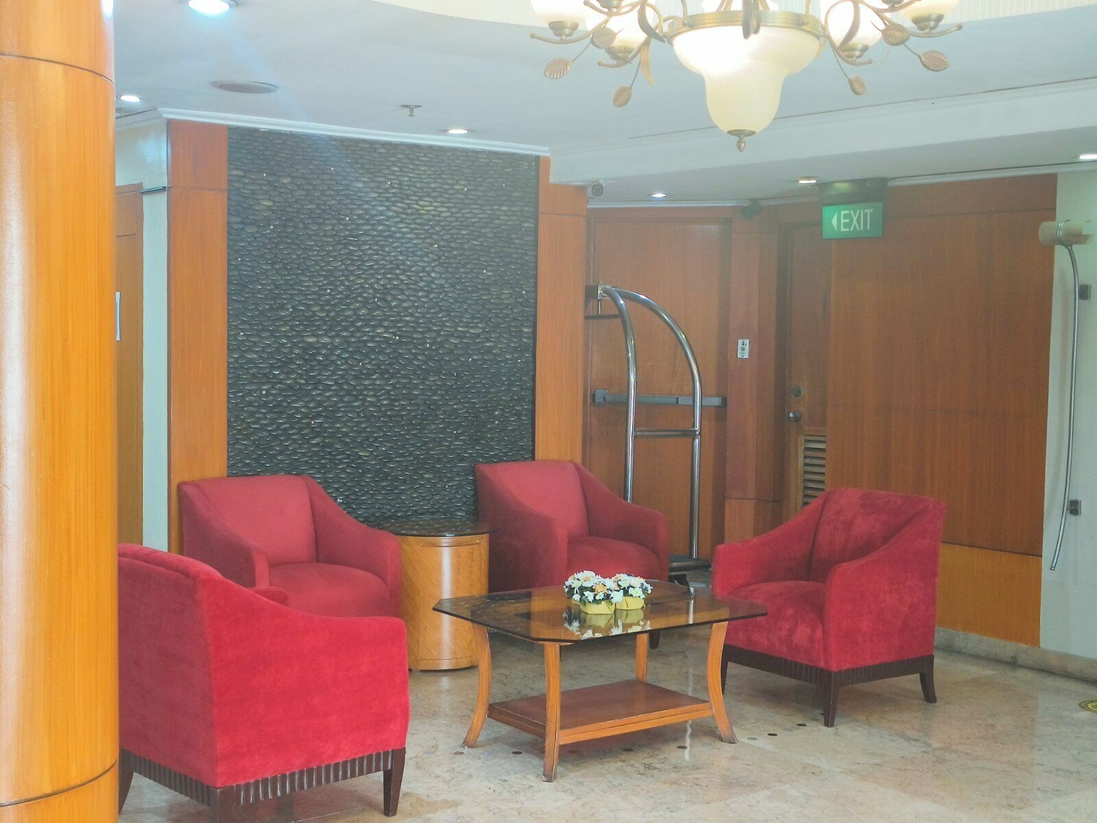 Um hall com cadeiras vermelhas, uma mesa de vidro com flores, uma parede texturizada e um sinal de saída.