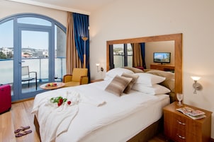 Quarto de hotel com uma cama grande, uma mesa de cabeceira de madeira, uma televisão, varanda com vista para a cidade e um espelho grande.