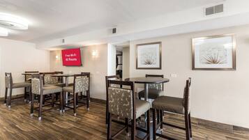 Quarto de hotel com área de refeições, piso de madeira, cadeiras marrom-escuro com almofadas estampadas e um letreiro vermelho indicando acesso gratuito à internet.