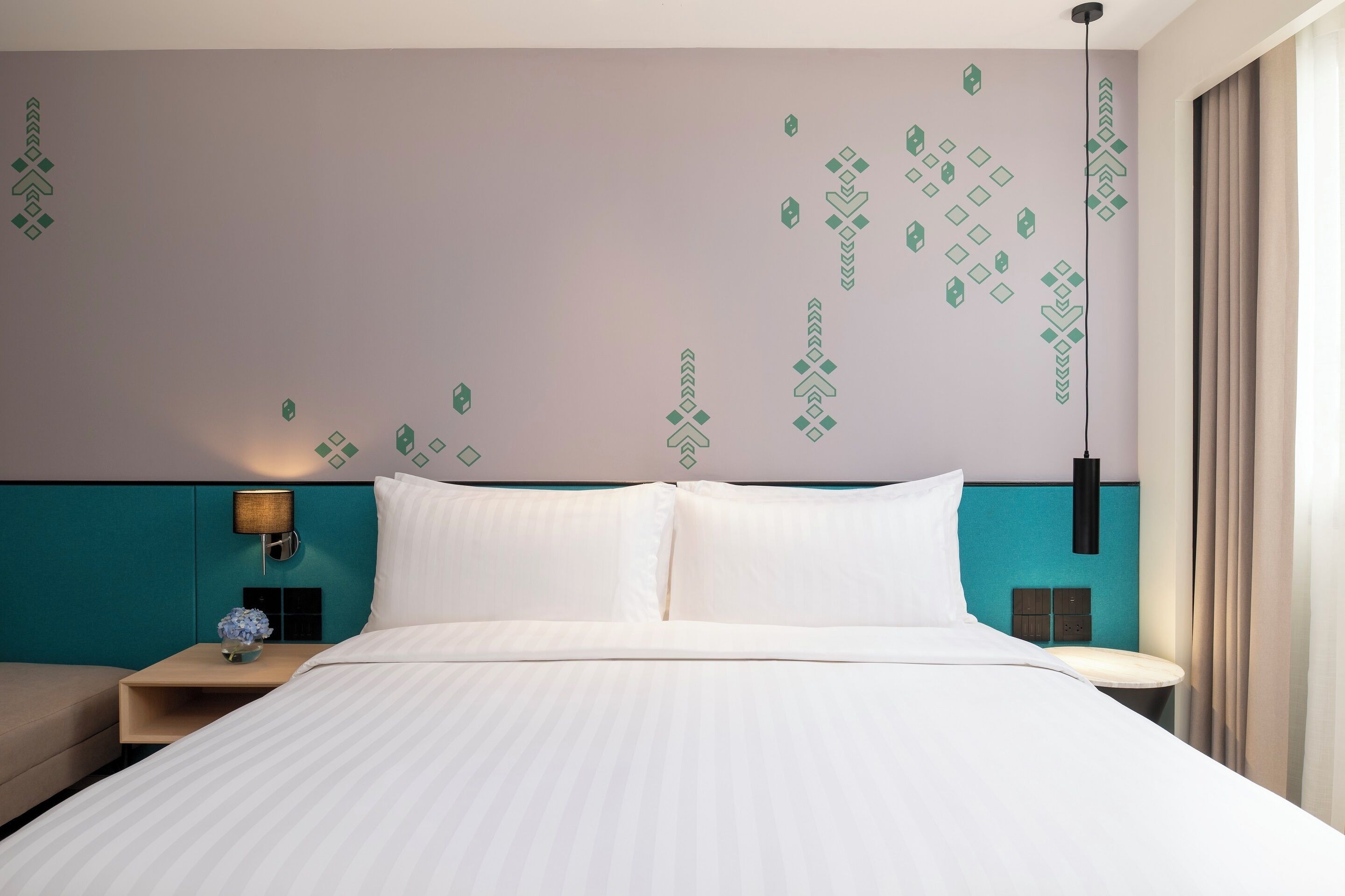 Quarto de hotel com cama, mesa de cabeceira com abajur, luminária na parede e janela com cortinas.