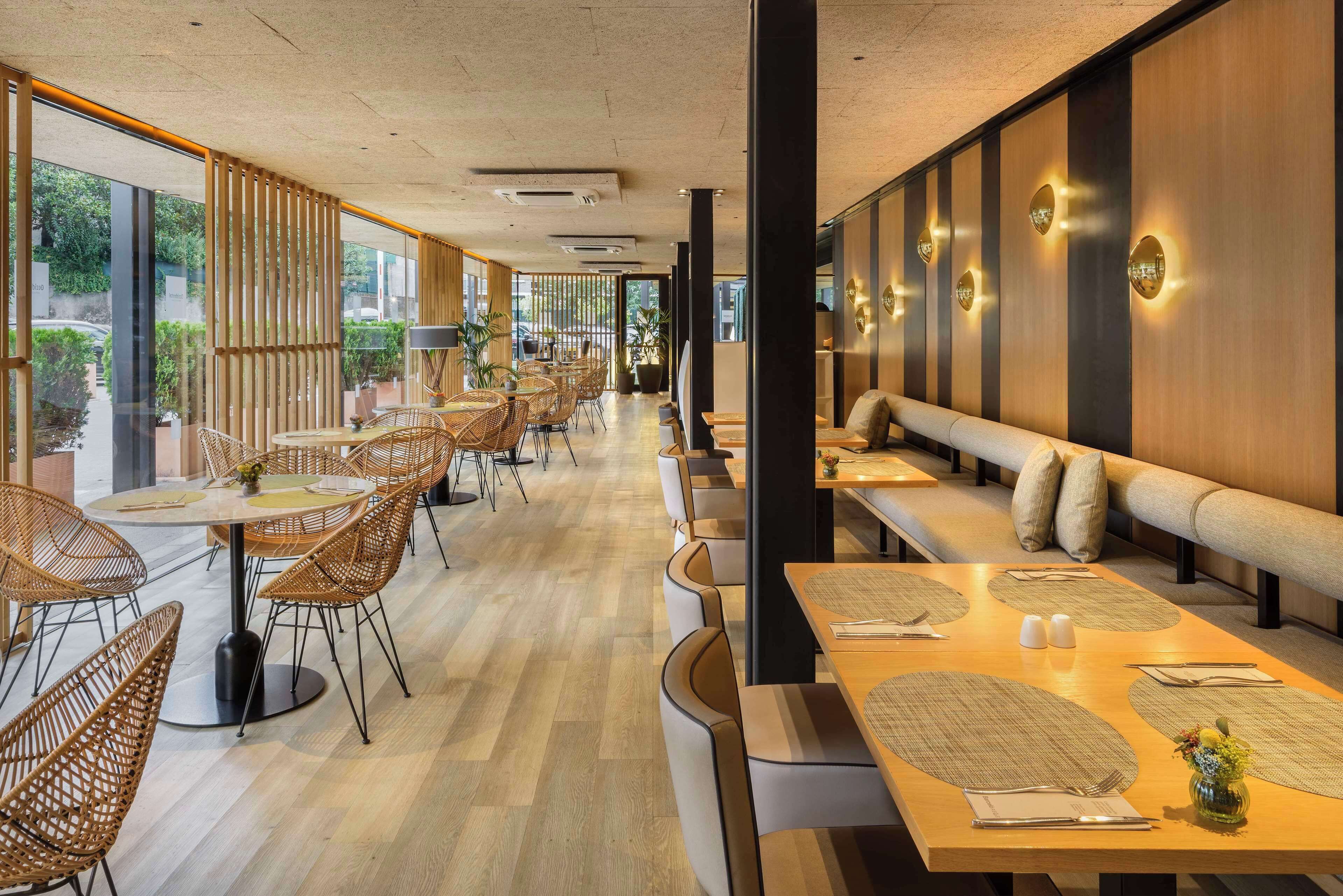 Um restaurante moderno com mesas e cadeiras de madeira, um balcão comprido com bancos e grandes janelas que oferecem vista para o exterior.