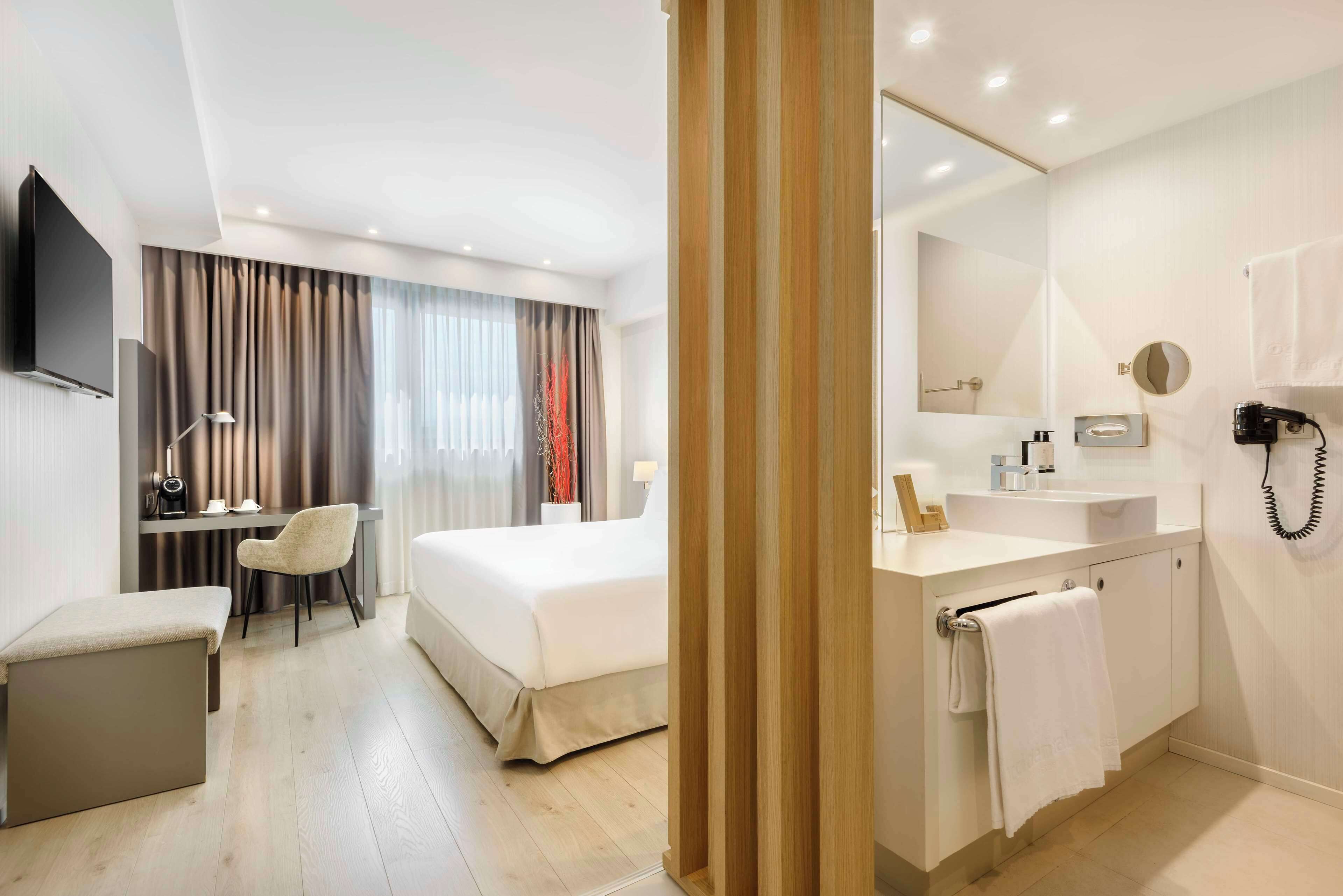 Quarto de hotel com cama, secretária com cadeira, televisão, candeeiro de mesa, espelho, lavatório e porta-toalhas.