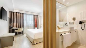 Quarto de hotel com cama, secretária com cadeira, televisão, candeeiro de mesa, espelho, lavatório e porta-toalhas.