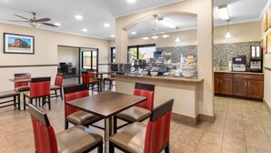 Uma sala de jantar de hotel com mesas e cadeiras, um balcão de cozinha com alimentos e um ventilador de teto.