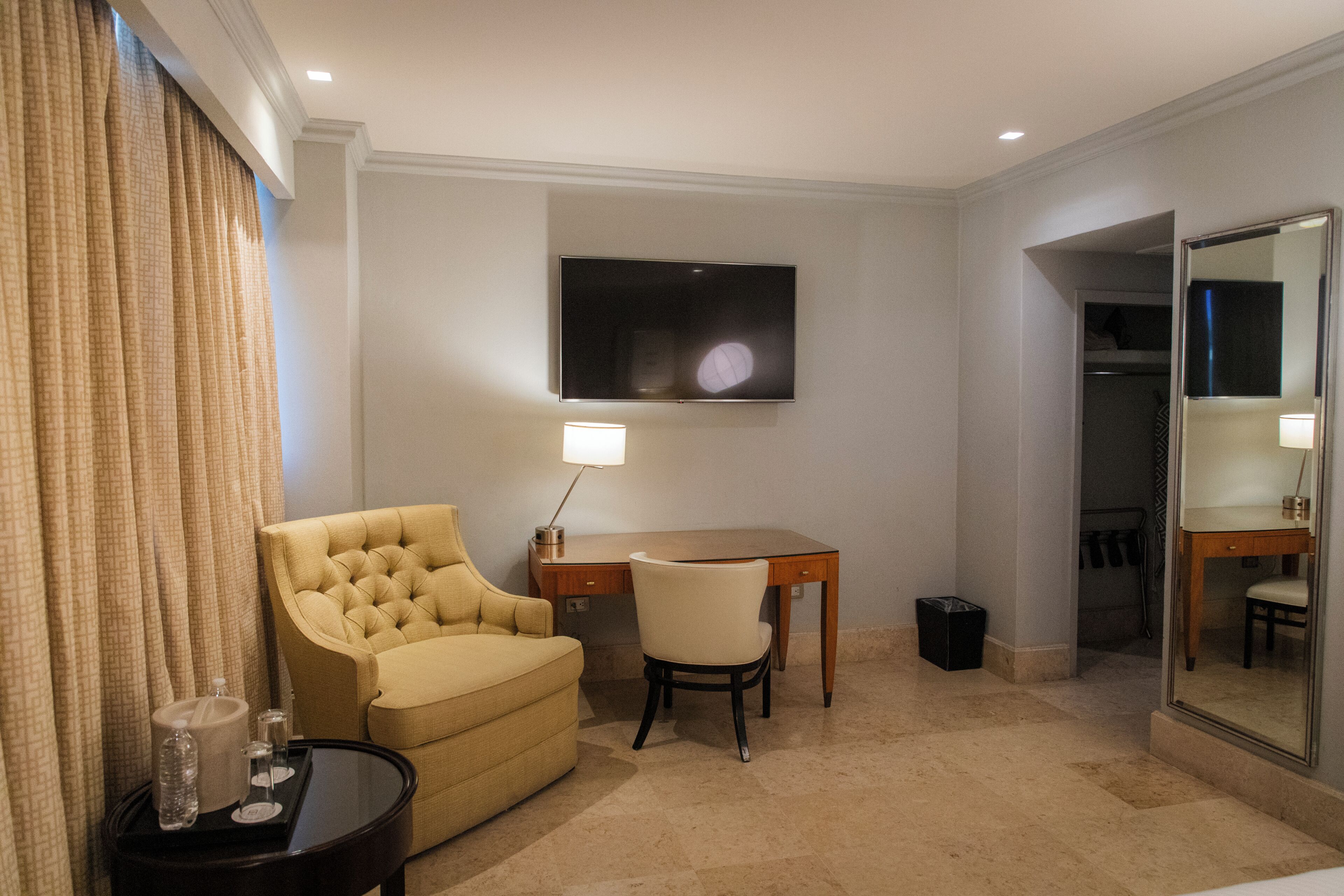 Quarto de hotel com poltrona estofada, escrivaninha com luminária, televisão de tela plana e um espelho grande.