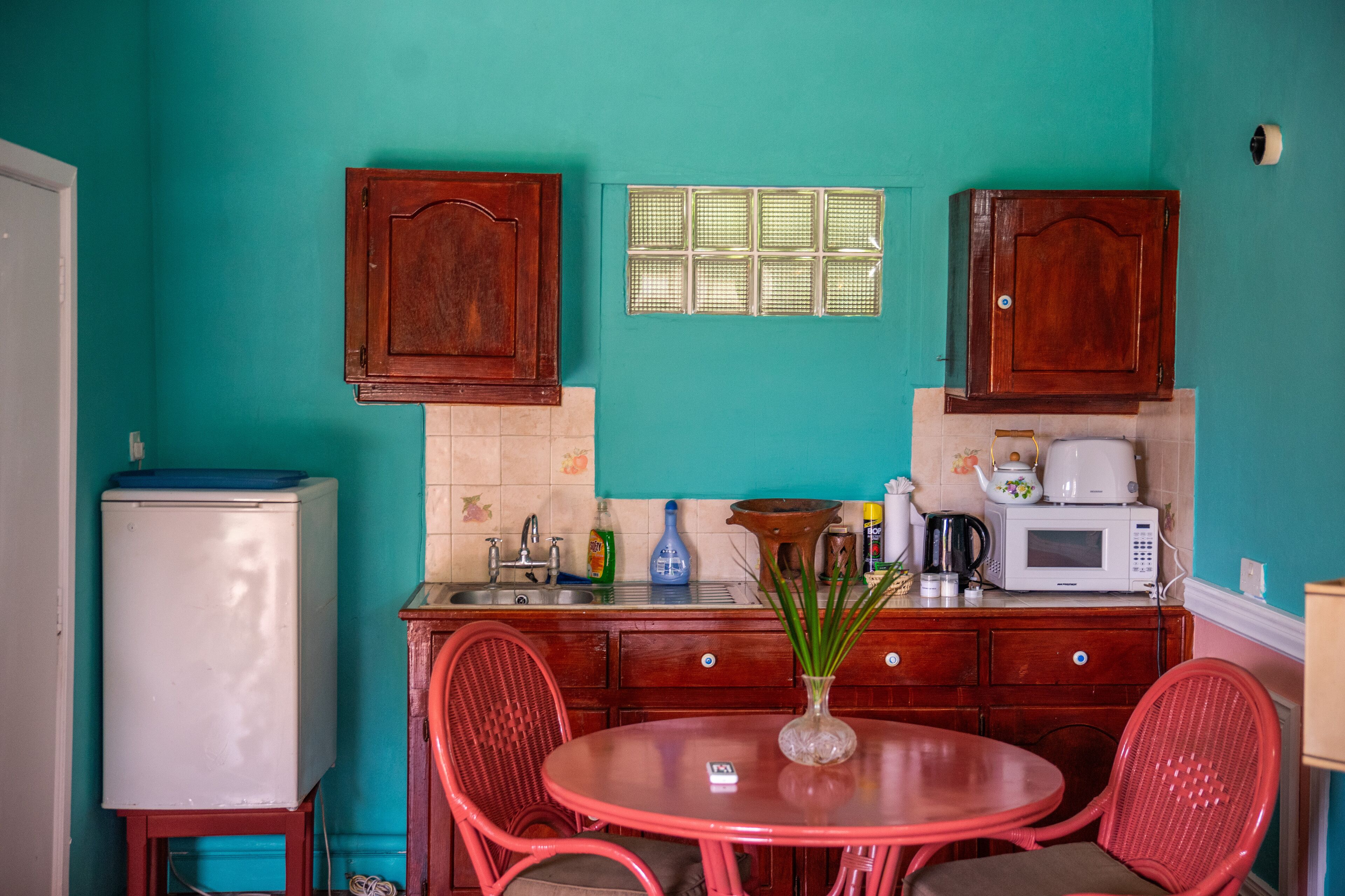 Uma cozinha compacta com paredes verde-água, armários de madeira, uma geladeira branca, micro-ondas e uma pequena mesa de jantar com duas cadeiras.
