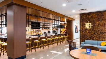 Uma área de bar moderna com um balcão comprido, vários banquetes e um espaço de estar com uma mesa redonda e cadeiras.