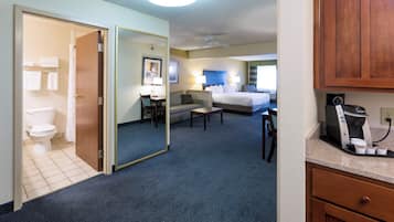 Quarto de hotel com banheiro, cama, escrivaninha, cadeira, cafeteira e espelho.