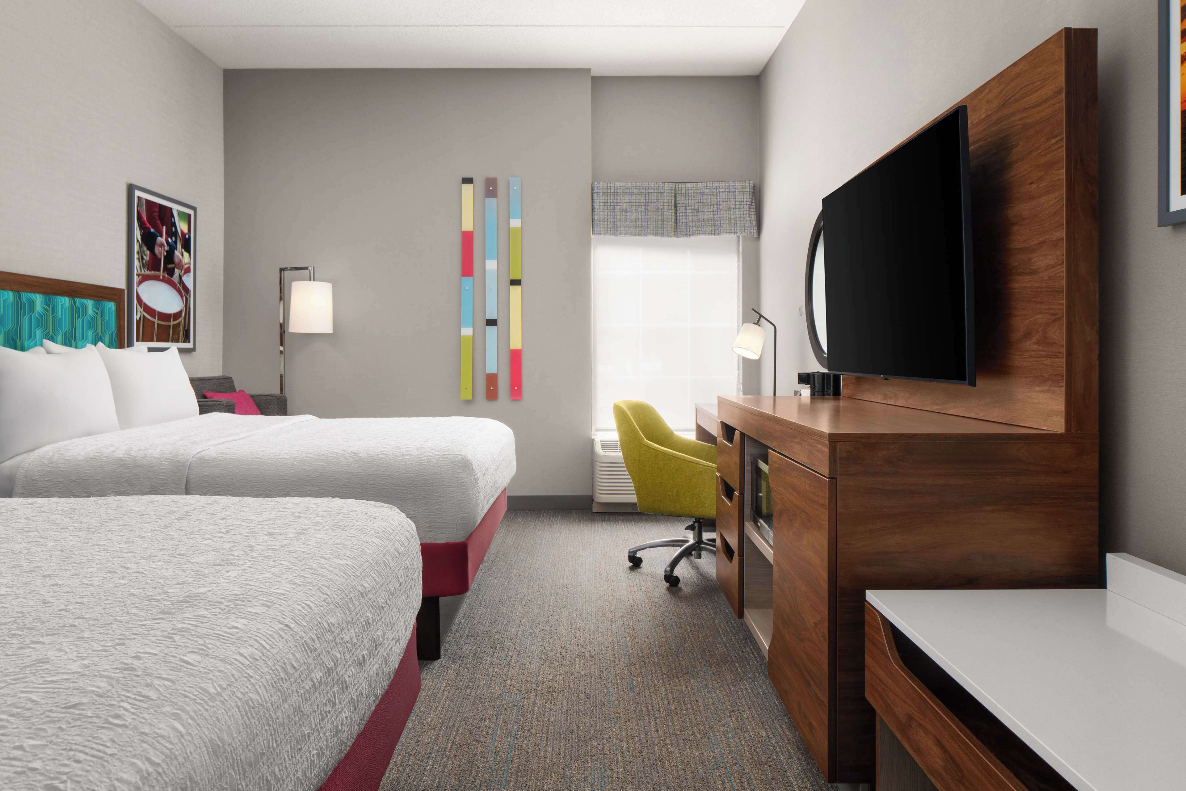 Quarto de hotel com duas camas, uma mesa de trabalho, televisão de tela plana, uma cadeira e uma janela com cortinas.