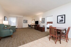 Quarto de hotel com área de estar, mesa de jantar com cadeiras, televisão, escrivaninha e cadeira.