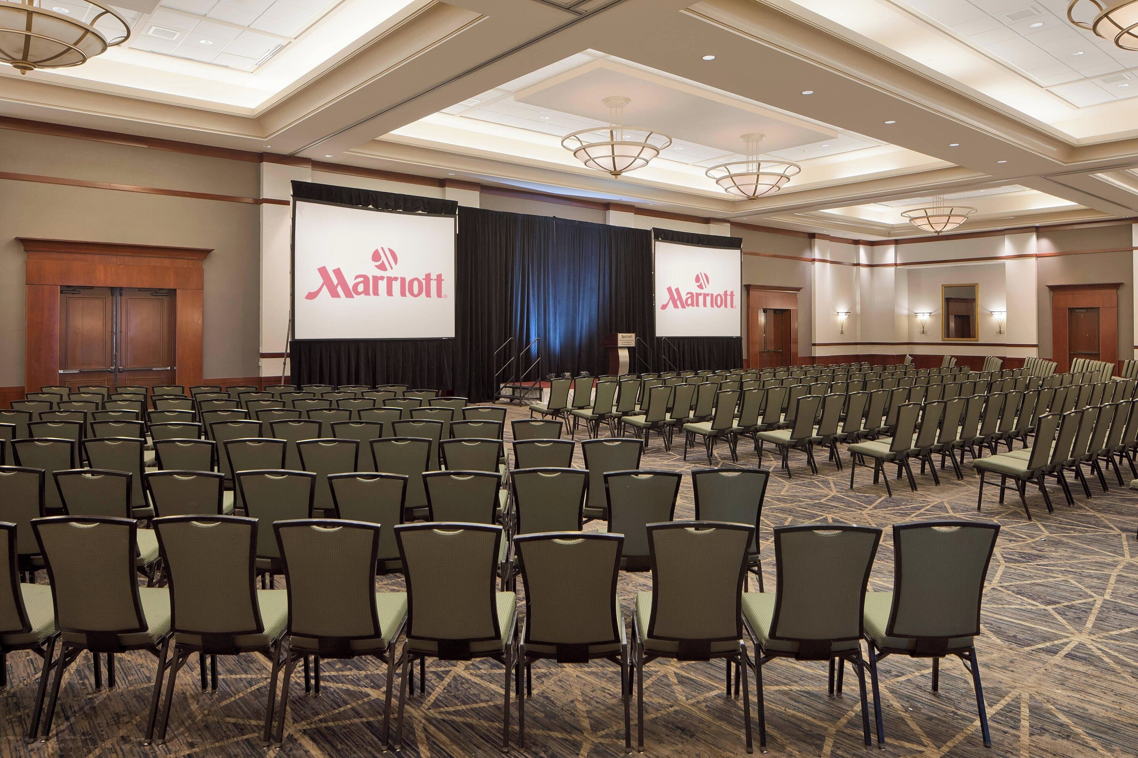 Uma grande sala de conferências com fileiras de cadeiras, um palco com um banner da Marriott e piso coberto de carpete.