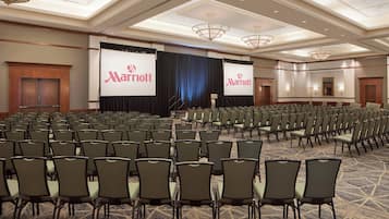 Uma grande sala de conferências com fileiras de cadeiras, um palco com um banner da Marriott e piso coberto de carpete.