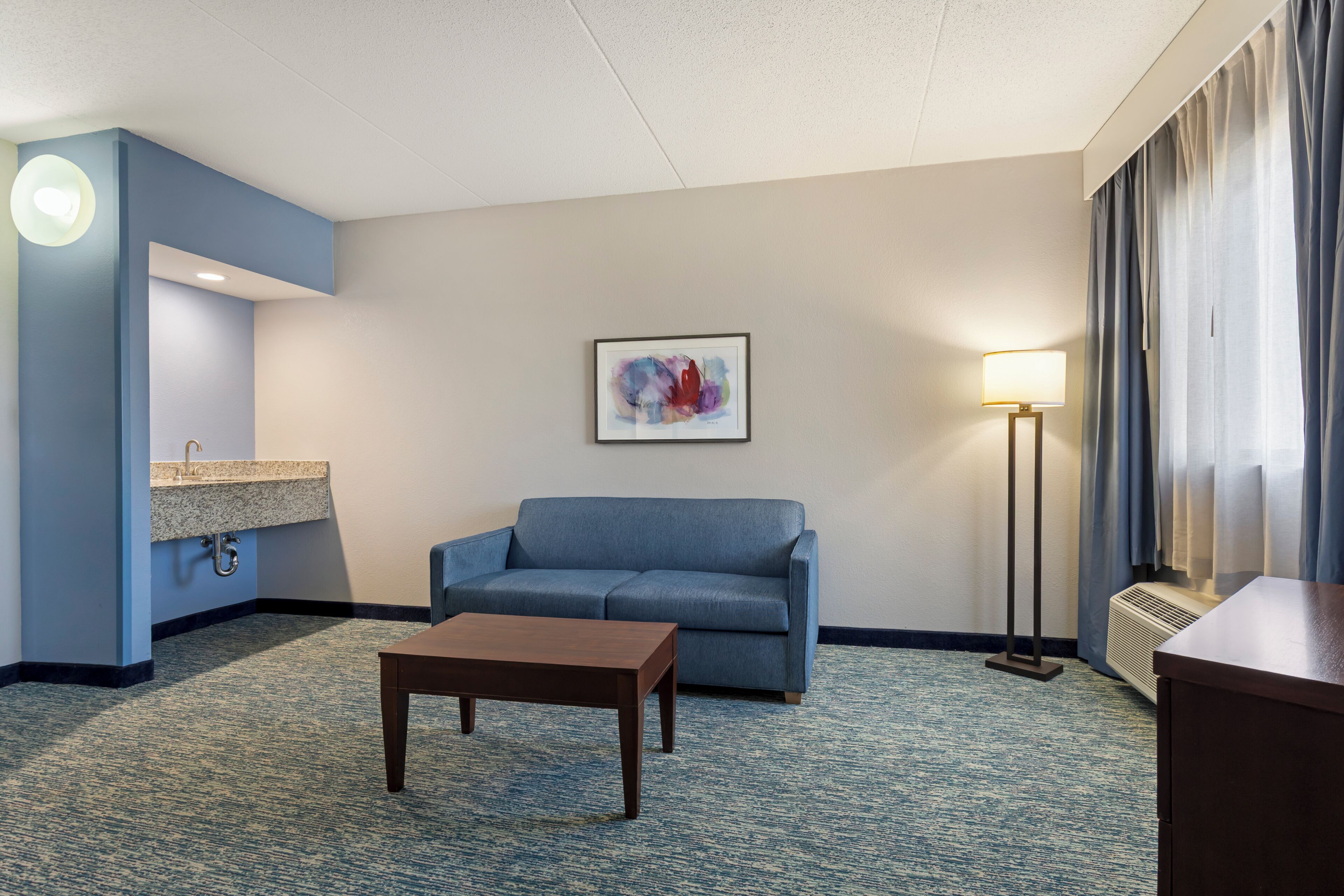 Quarto de hotel com sofá azul, uma mesinha, um quadro na parede, uma luminária e uma janela com cortinas.