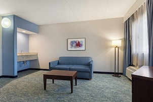 Quarto de hotel com sofá azul, uma mesinha, um quadro na parede, uma luminária e uma janela com cortinas.