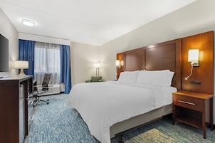 Quarto de hotel com cama, escrivaninha, cadeira, mesa de cabeceira e abajur.
