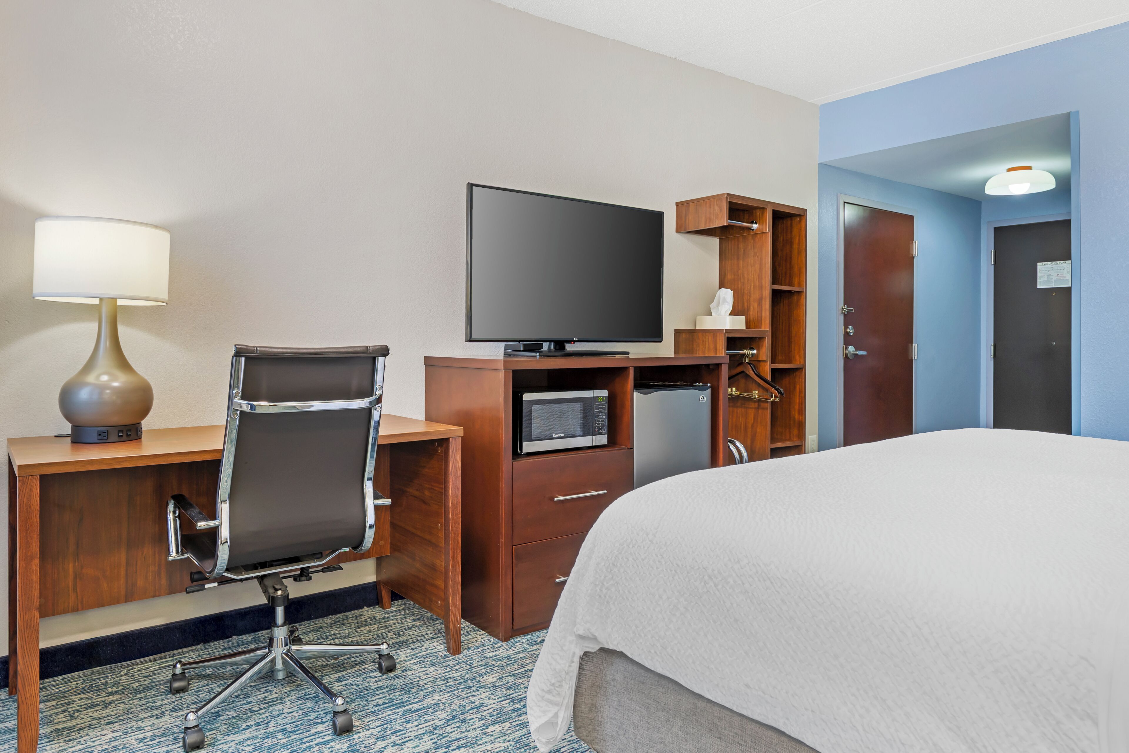 Quarto de hotel com cama, escrivaninha, cadeira, televisão e minibar.