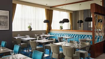 Uma sala de jantar de hotel com mesas e cadeiras, um banco acolchoado azul e janelas amplas com cortinas.